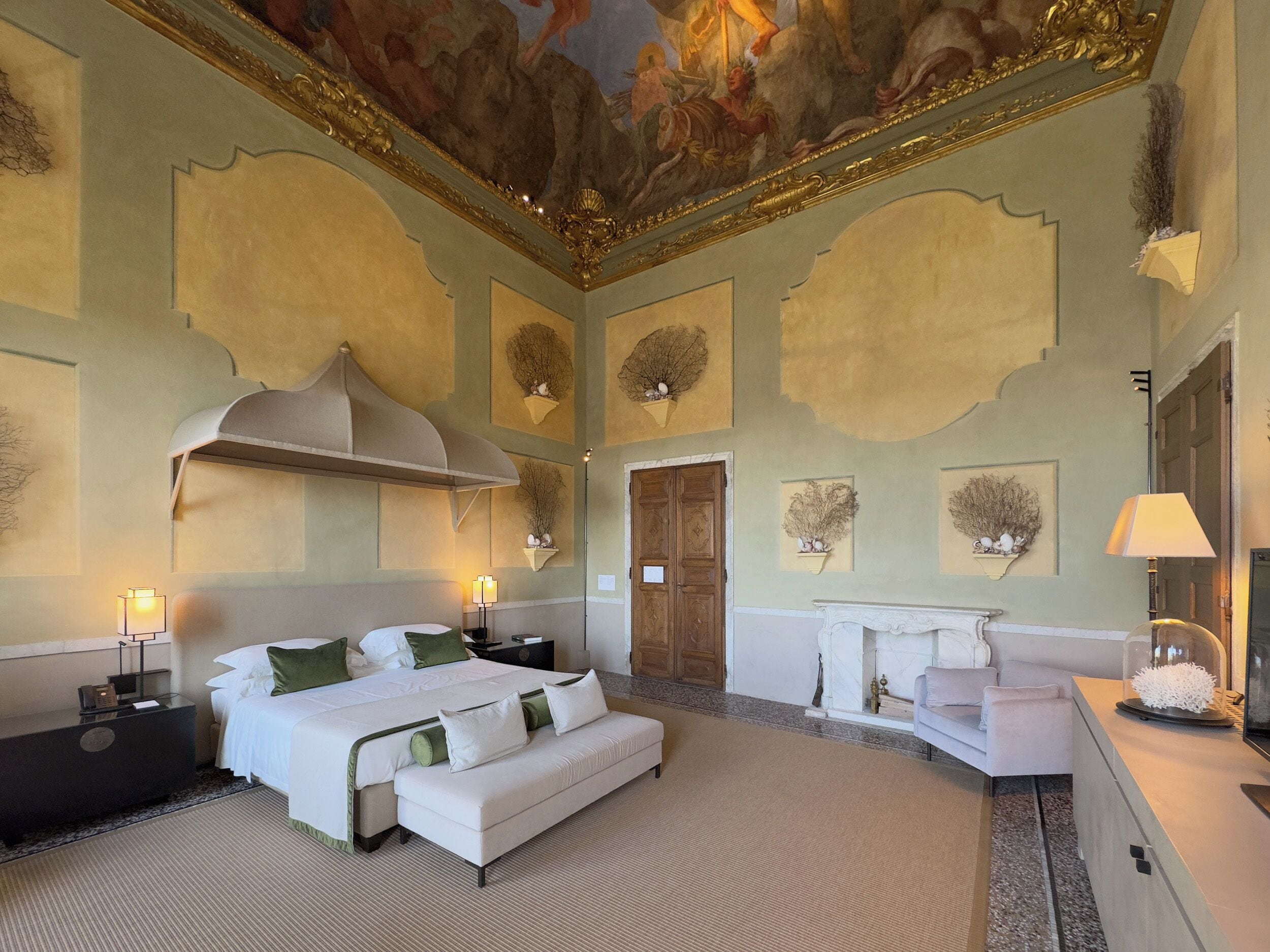 Quarto de hotel com cama grande, escrivaninha, cadeira, lareira e um mural no teto.
