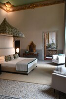 Quarto de hotel com uma cama grande, área de estar, espelho e teto decorativo.