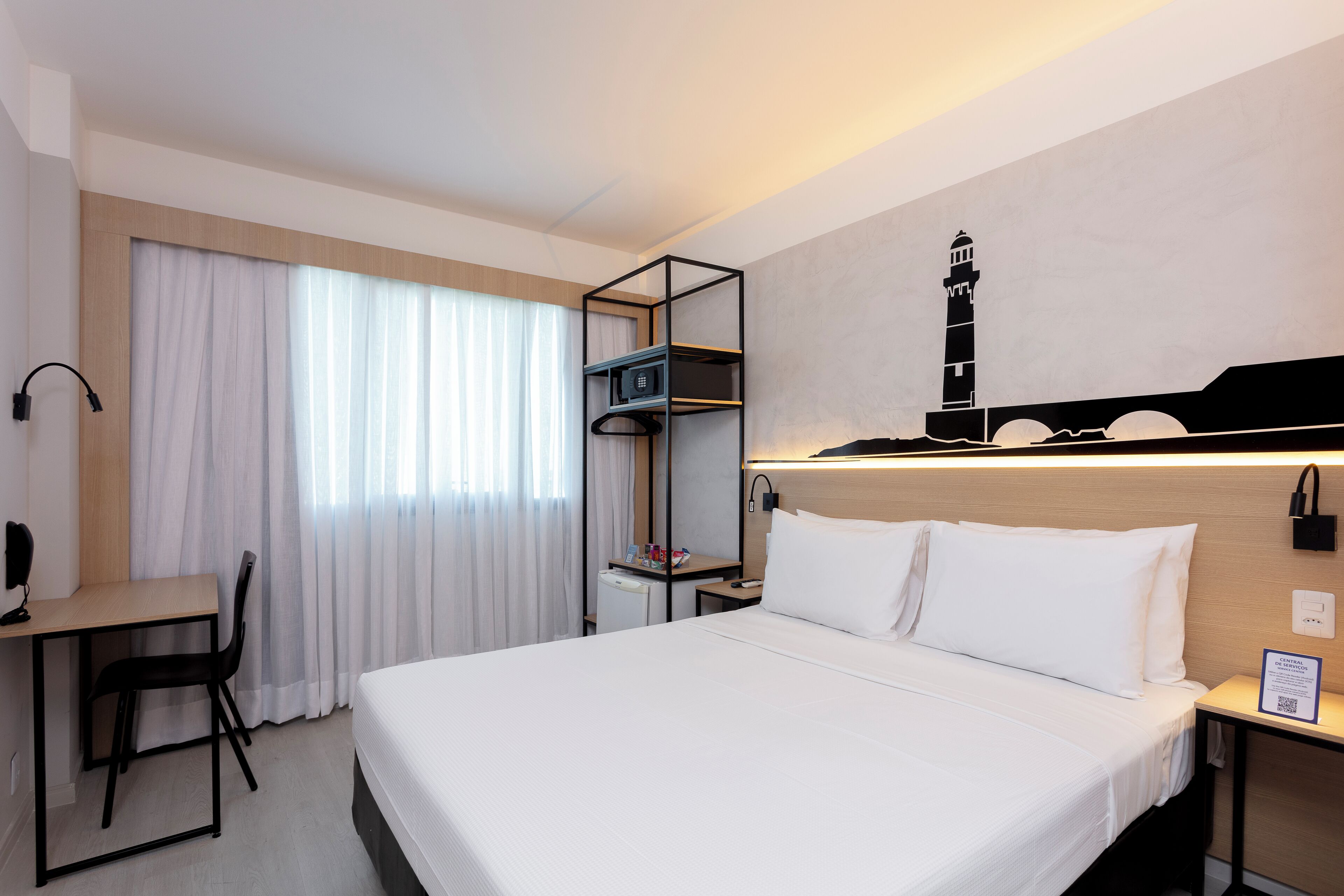 Quarto de hotel com cama, escrivaninha, cadeira e prateleira fixada na parede.