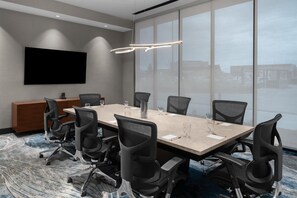 Sala de conferências com uma mesa comprida, cadeiras, uma tela de TV e um janelão.