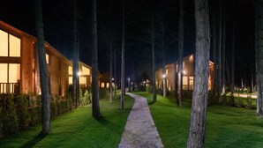Uma trilha leva a uma fileira de edifícios de madeira com janelas iluminadas, cercada por árvores e grama bem cuidada.