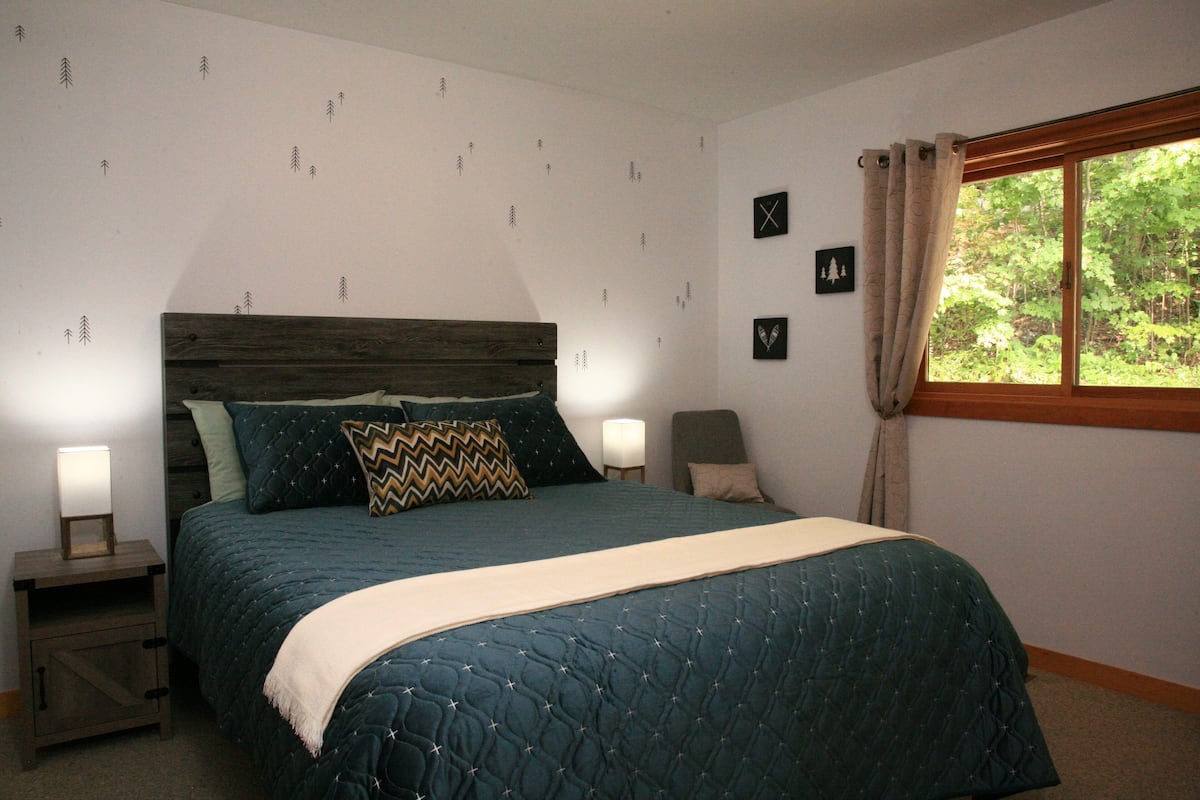 Quarto com uma cama grande, duas mesas de cabeceira, uma janela com cortinas e decorações nas paredes.