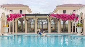 Uma piscina com uma pessoa sentada na borda, cercada por arcos e trepadeiras cor-de-rosa com flores.