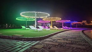 Área externa à noite com cadeiras de descanso e guarda-sóis iluminados.