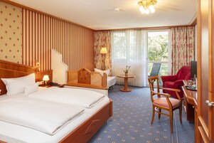 Quarto de hotel com uma cama grande, uma escrivaninha com cadeira, uma mesinha e uma varanda com cadeira.