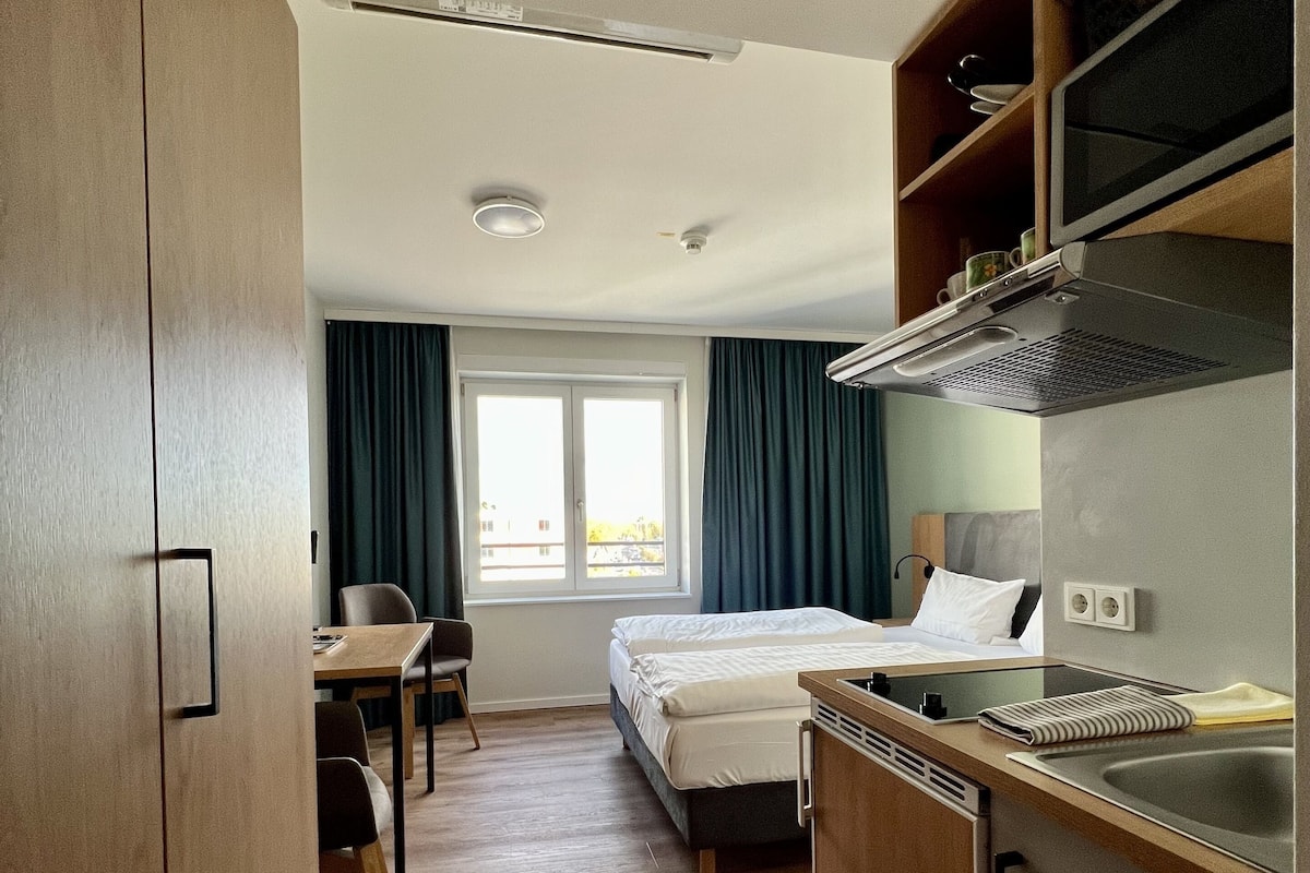Quarto de hotel compacto com cozinha compacta, cama, escrivaninha e janela com cortinas.