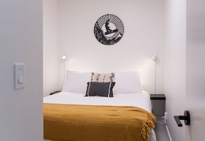 Cama bem arrumada com manta cor mostarda, almofadas brancas e almofada estampada em preto e branco. Uma arte na parede circular retrata um snowboarder.