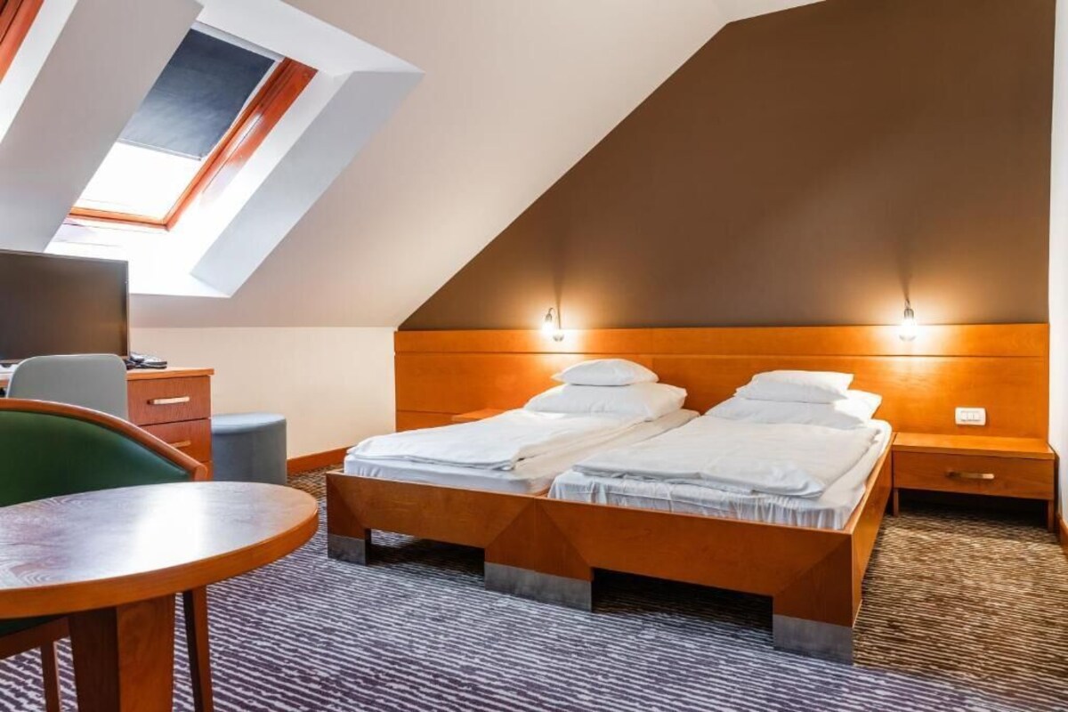 Quarto de hotel com teto inclinado, cama com roupa de cama branca, escrivaninha com cadeira e uma pequena mesa.