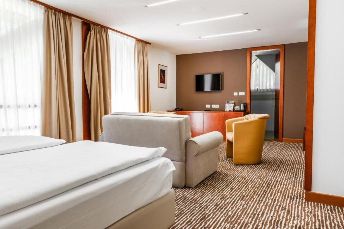 Quarto de hotel com uma cama, duas poltronas, uma escrivaninha, televisão e um tapete estampado.