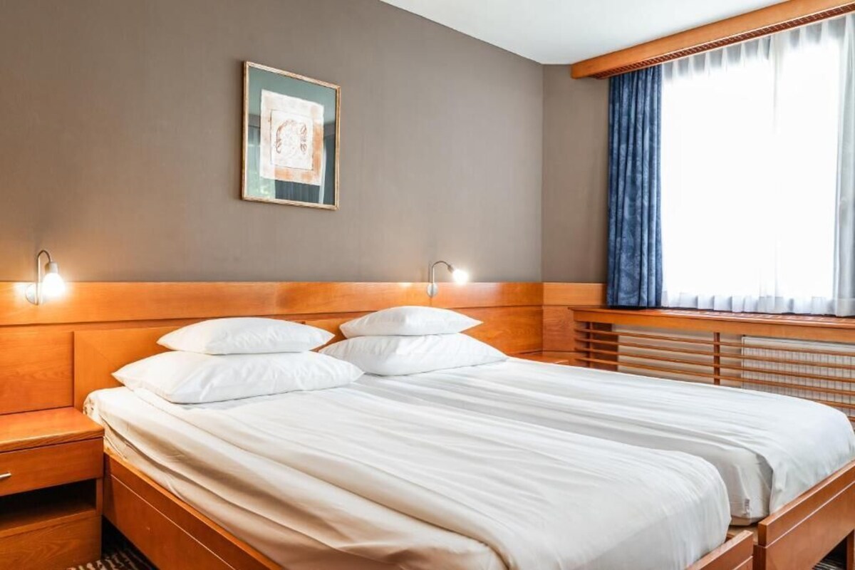 Quarto de hotel com uma cama grande, mesas de cabeceira, painel de cabeceira, uma janela com cortinas e um quadro emoldurado na parede.