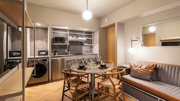 Uma cozinha compacta com eletrodomésticos modernos, uma sala de jantar com mesa e cadeiras redondas e uma área de estar com sofá e almofadas.