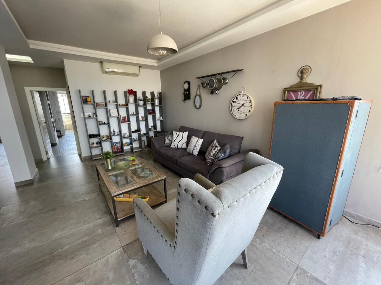 Uma sala de estar com sofá, poltrona, mesa de centro e prateleiras com objetos decorativos.