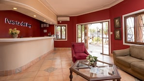 Uma área de recepção com parede vermelha, balcão de recepção, poltrona vermelha, mesa de centro de vidro com um vaso e sofá bege.