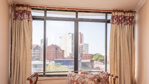 Quarto com sofá, mesa de vidro, janelas amplas com cortinas e vista para a cidade.