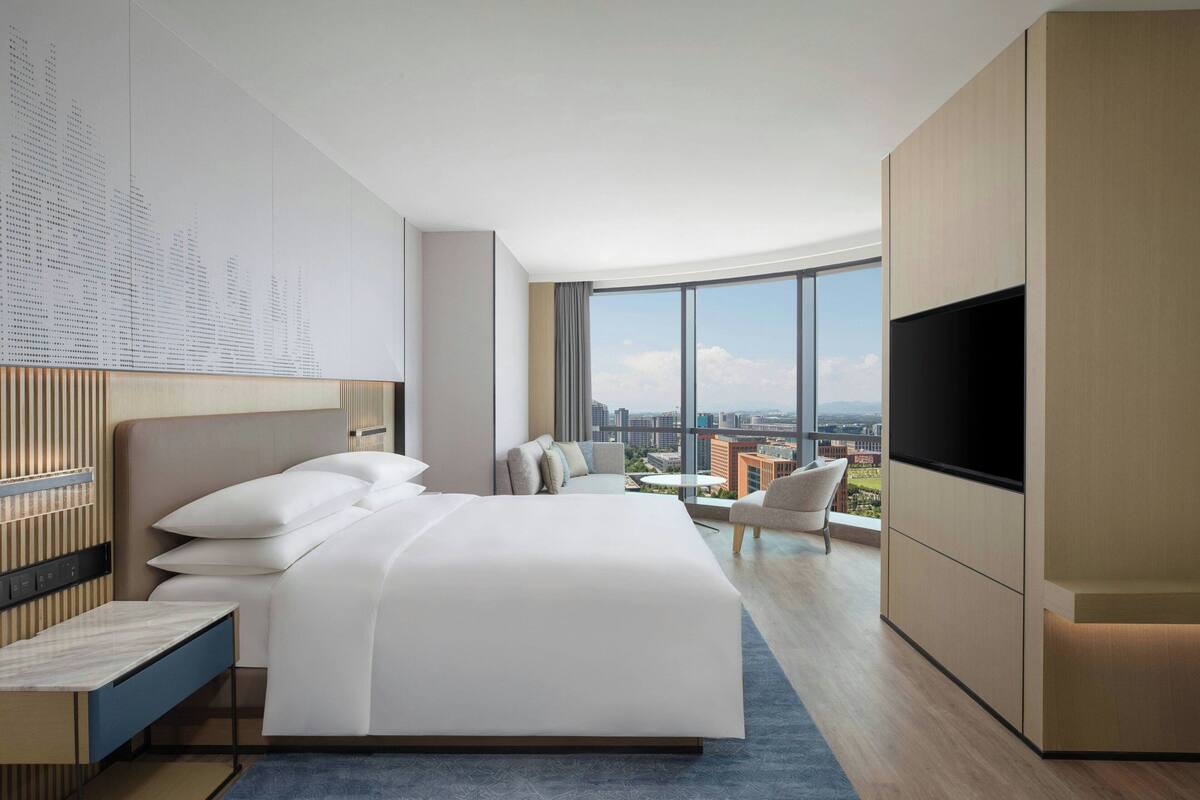 Quarto de hotel moderno com uma cama grande, televisão de tela plana, vista para a cidade e área de estar com vista panorâmica do horizonte urbano.