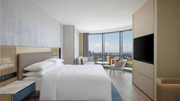 Quarto de hotel moderno com uma cama grande, televisão de tela plana, vista para a cidade e área de estar com vista panorâmica do horizonte urbano.