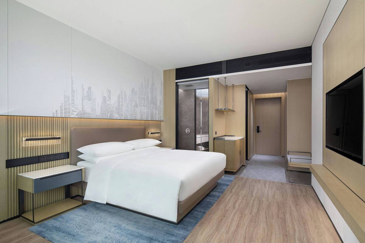 Quarto de hotel moderno com uma cama grande, uma escrivaninha, uma televisão e uma porta deslizante para o banheiro.