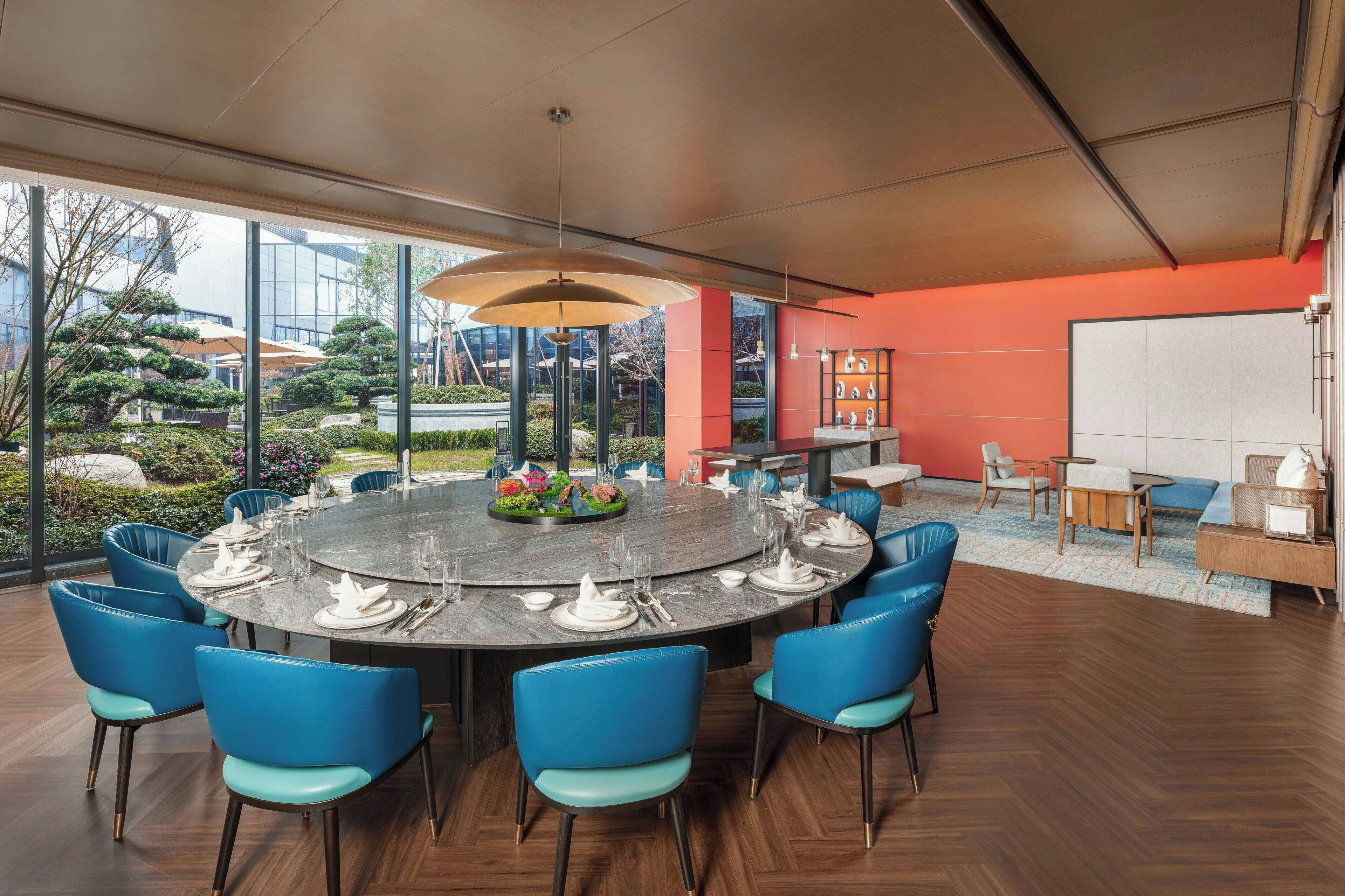 Uma área de jantar moderna com uma mesa redonda posta para seis pessoas, rodeada por cadeiras azuis, uma grande janela com vista para o exterior e uma parede de destaque vermelha.