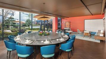 Uma área de jantar moderna com uma mesa redonda posta para seis pessoas, rodeada por cadeiras azuis, uma grande janela com vista para o exterior e uma parede de destaque vermelha.
