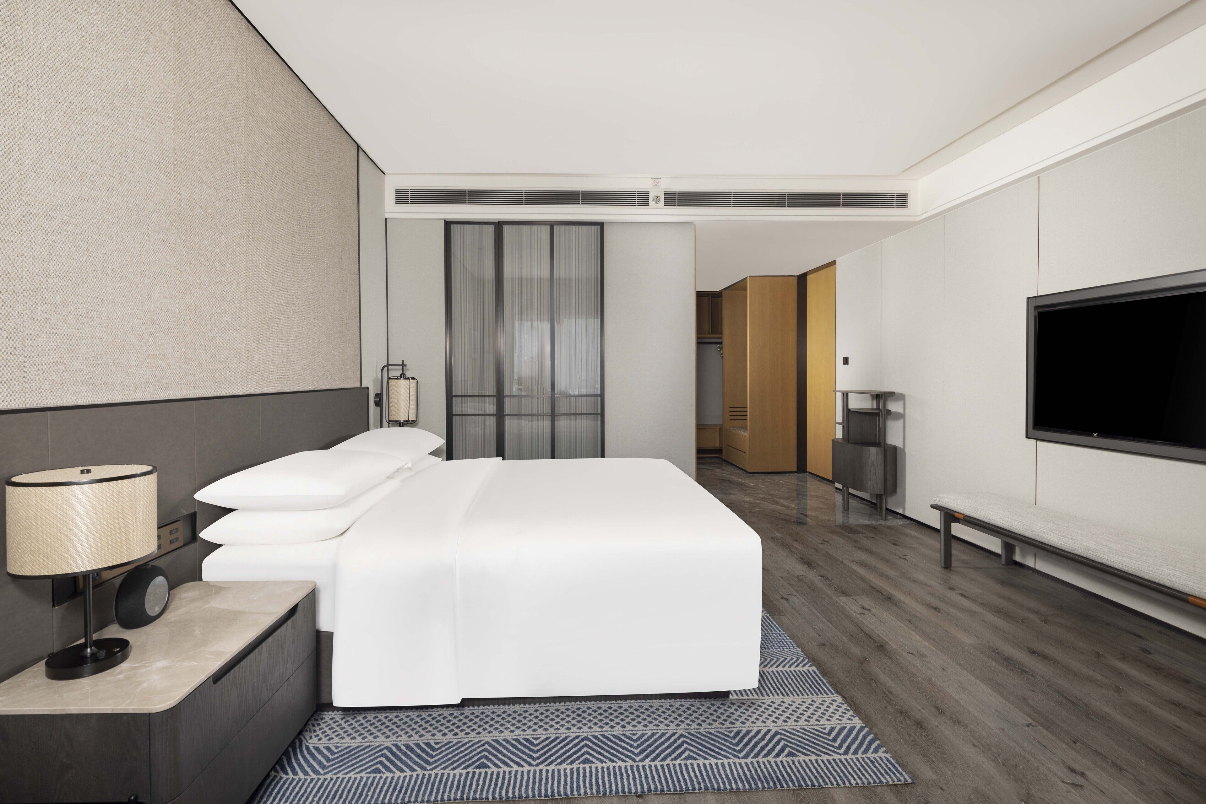 Quarto de hotel moderno com uma cama grande, televisão de tela plana, mesa de cabeceira com abajur e vista para o corredor.
