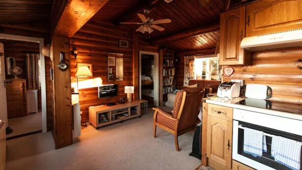 Interior acolhedor de cabana com teto de madeira, ventilador de teto, televisão, mesa de jantar e cozinha com fogão.