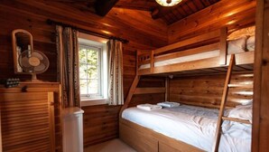 Quarto de cabana de madeira com beliches, uma pequena escrivaninha, um espelho e uma janela com cortinas.