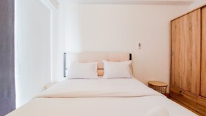 Uma cama bem arrumada com lençóis e almofadas brancos, um guarda-roupa de madeira e uma pequena mesa redonda ao lado da cama.