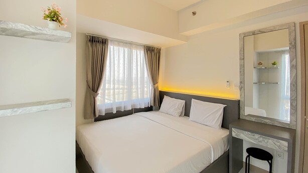 Quarto de hotel com cama, escrivaninha, cadeira, espelho e prateleira com uma planta.