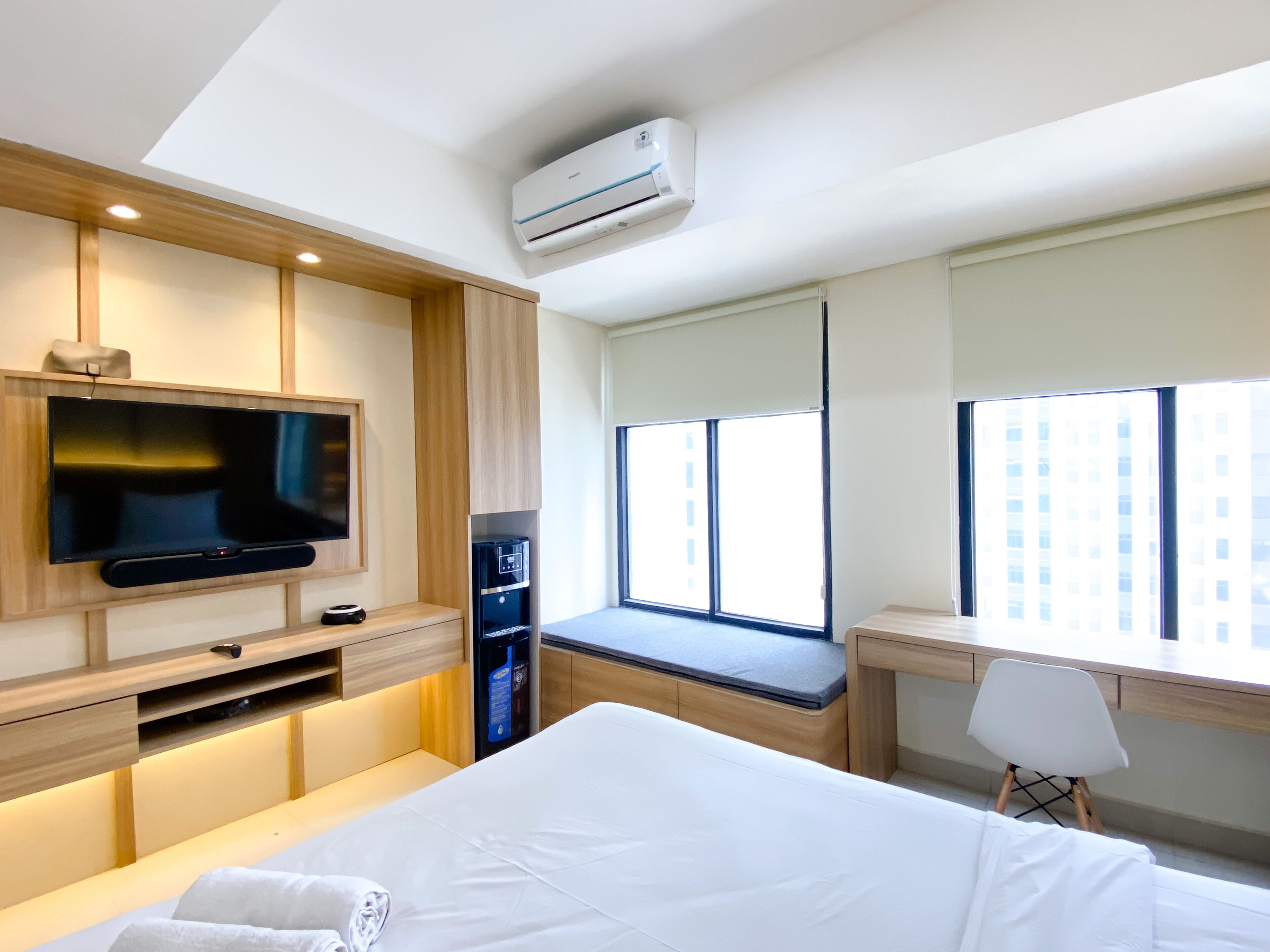 Quarto de hotel com cama, mesa de trabalho, televisão, dispensador de água e janela com vista para edifícios.