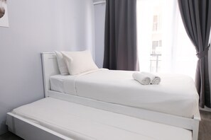 Uma cama de solteiro com roupa de cama e almofadas brancas, uma pequena mesa de cabeceira e uma janela com cortinas.