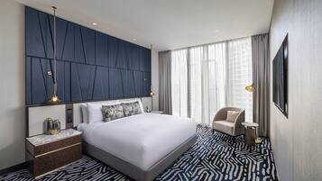 Quarto de hotel moderno com uma cama grande, mesa de cabeceira, poltrona e televisão.