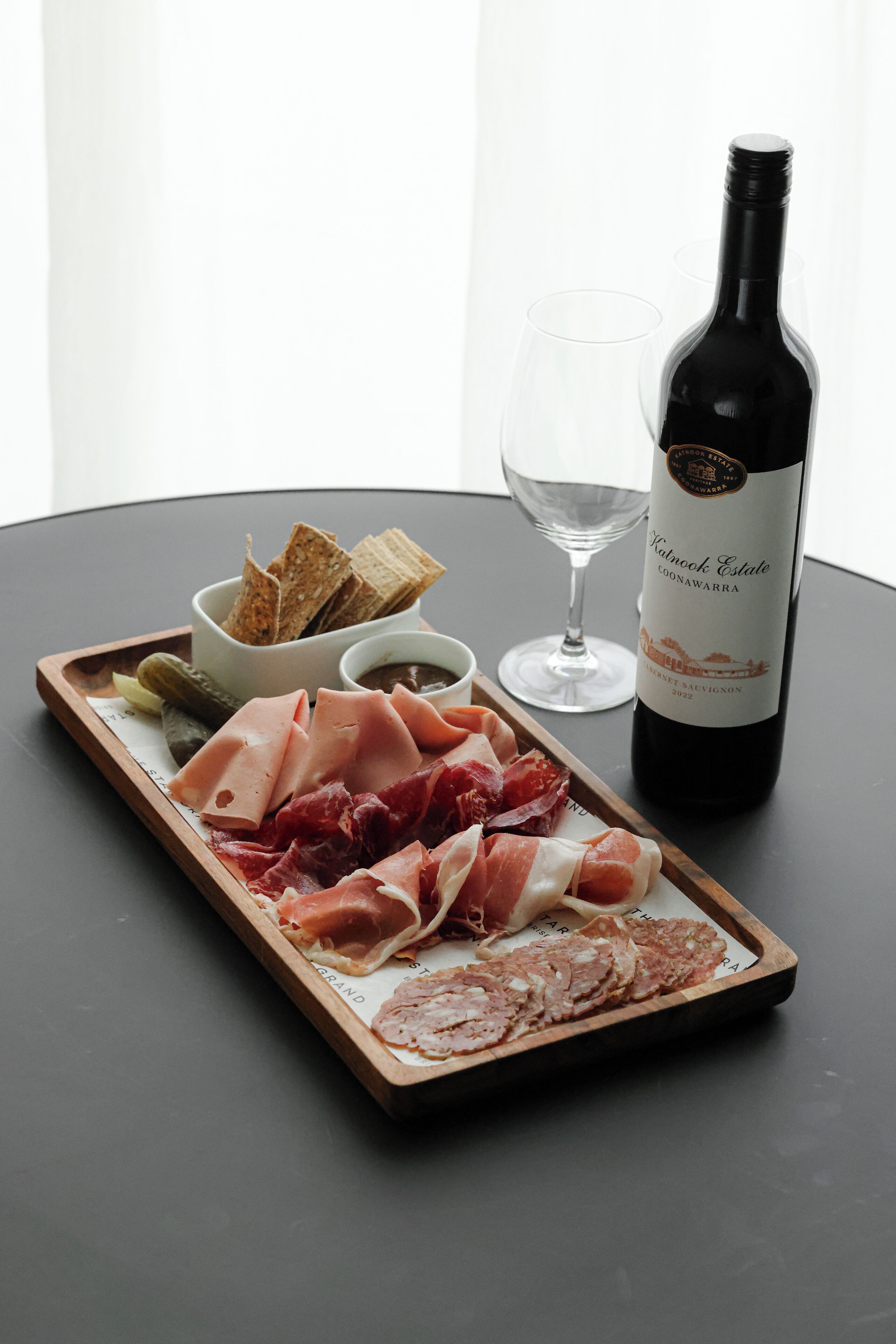 Uma tábua de madeira com diversos tipos de carne, uma garrafa de vinho e um copo sobre uma mesa.