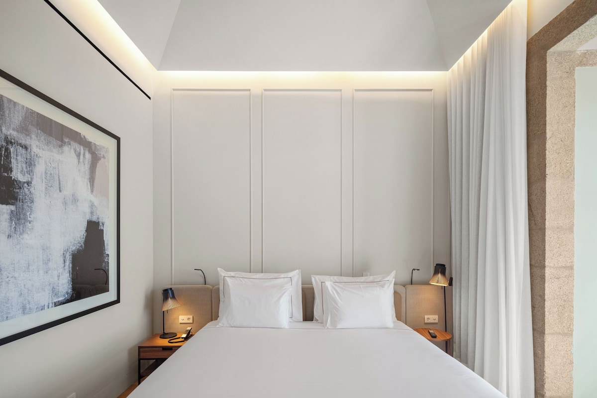 Quarto de hotel moderno com uma cama grande, duas mesas de cabeceira, uma obra de arte na parede e vista para o exterior.