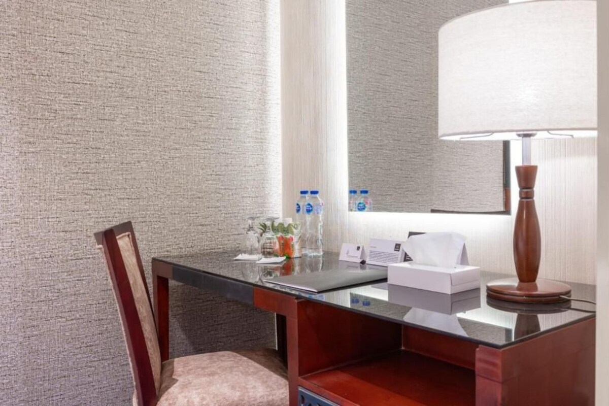 Quarto de hotel com secretária de madeira, cadeira, espelho, candeeiro e pequena mesa com artigos de higiene pessoal.