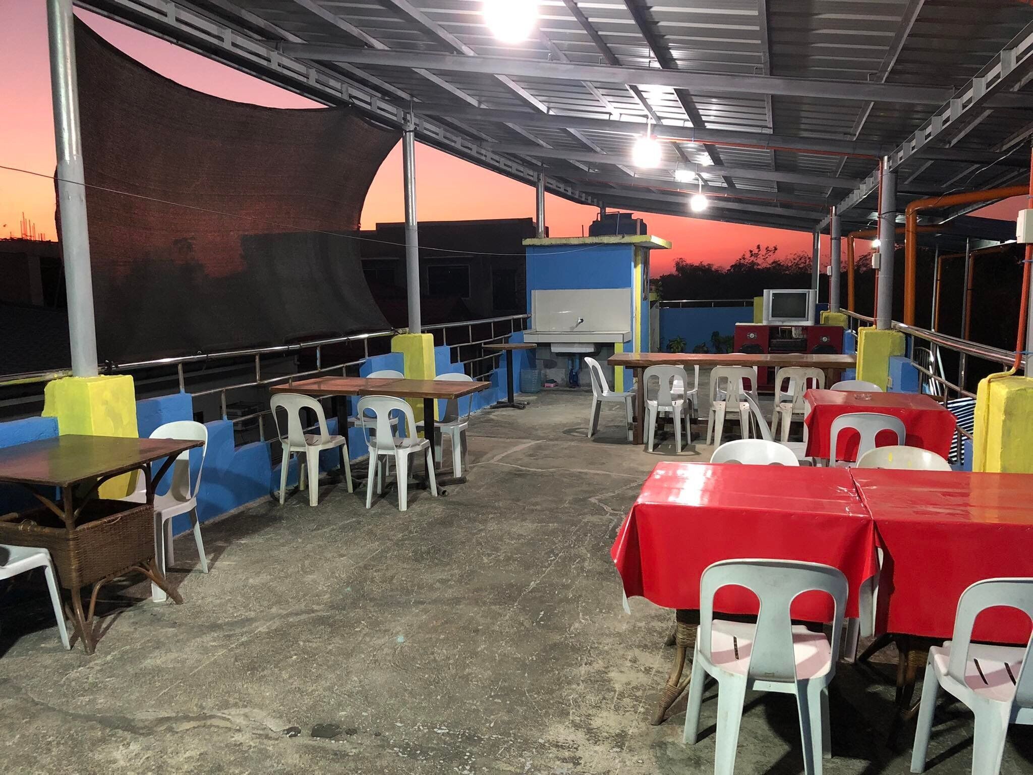 Uma área de refeições ao ar livre com mesas, cadeiras, uma tela grande e uma estrutura de telhado coberto.