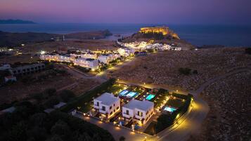 Um resort costeiro com villas, área de piscina e uma estrutura fortificada no topo de uma colina.