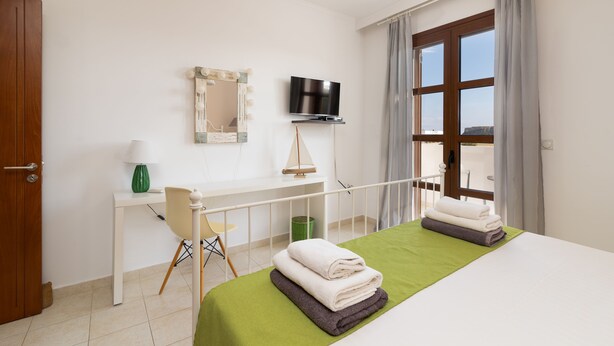 Quarto de hotel com cama, escrivaninha, cadeira, televisão e janela com cortinas.