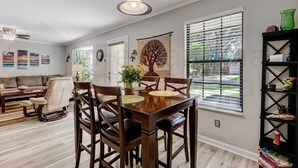 Uma sala de estar espaçosa com um sofá, uma mesa de jantar com cadeiras, um ventilador de teto e uma grande janela com persianas.
