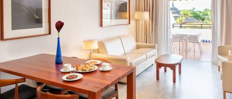 Quarto de hotel com mesa de jantar de madeira, um sofá, uma mesinha redonda e varanda com vista para vegetação e um edifício.