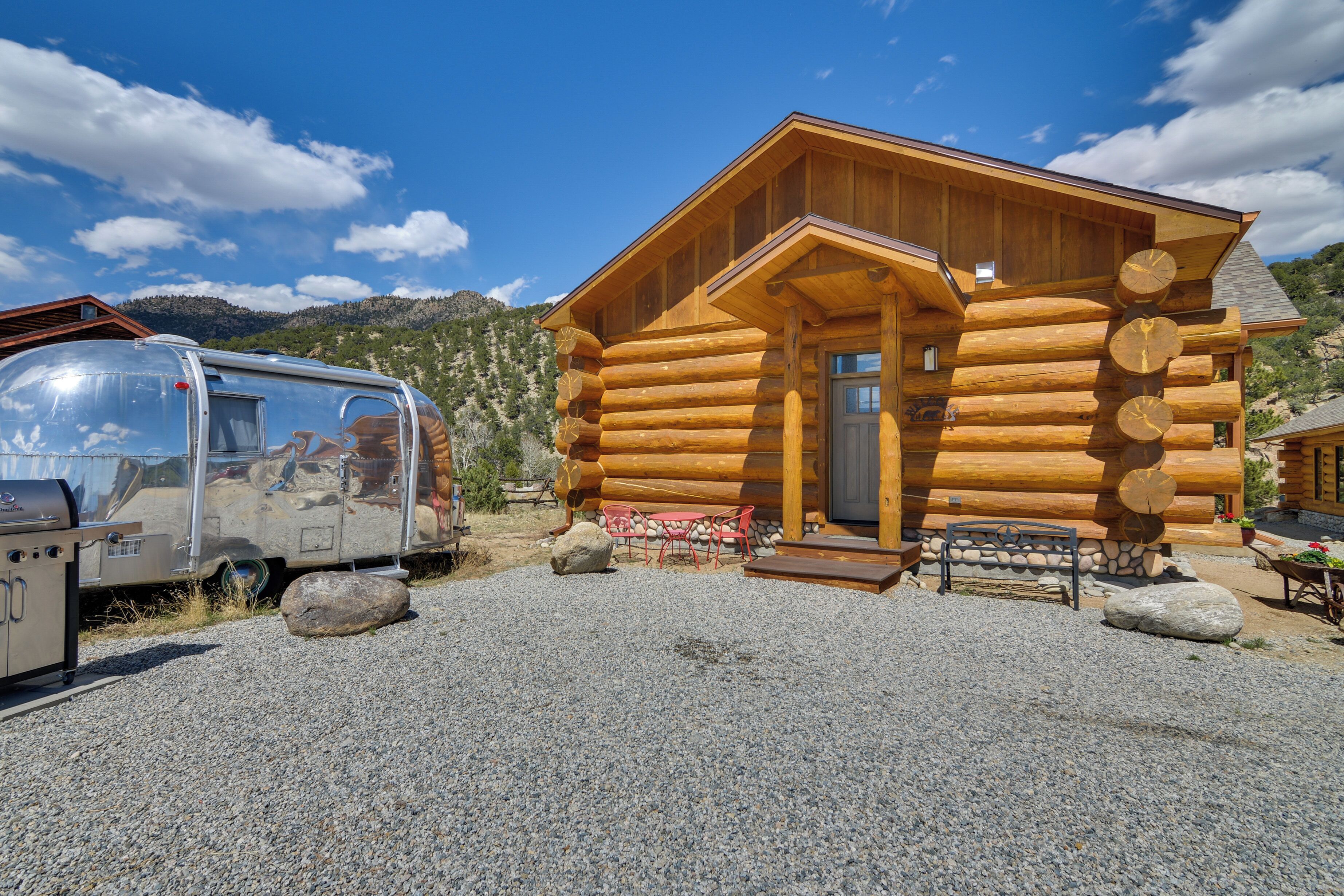 Uma cabana de tronco com um caminho de acesso de cascalho, uma van camper estacionada e montanhas ao fundo.