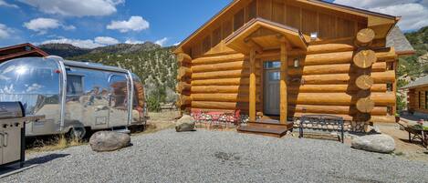 Uma cabana de tronco com um caminho de acesso de cascalho, uma van camper estacionada e montanhas ao fundo.