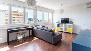 Uma sala de estar moderna com um sofá, uma escrivaninha e uma TV. Janelas amplas proporcionam vista para prédios lá fora.