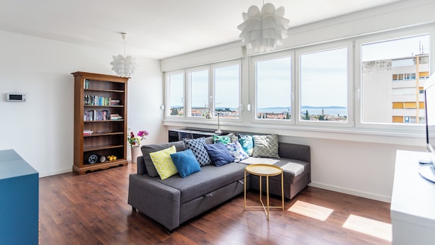 Uma sala de estar moderna com um sofá cinzento, uma estante de madeira e amplas janelas com vista para a cidade.