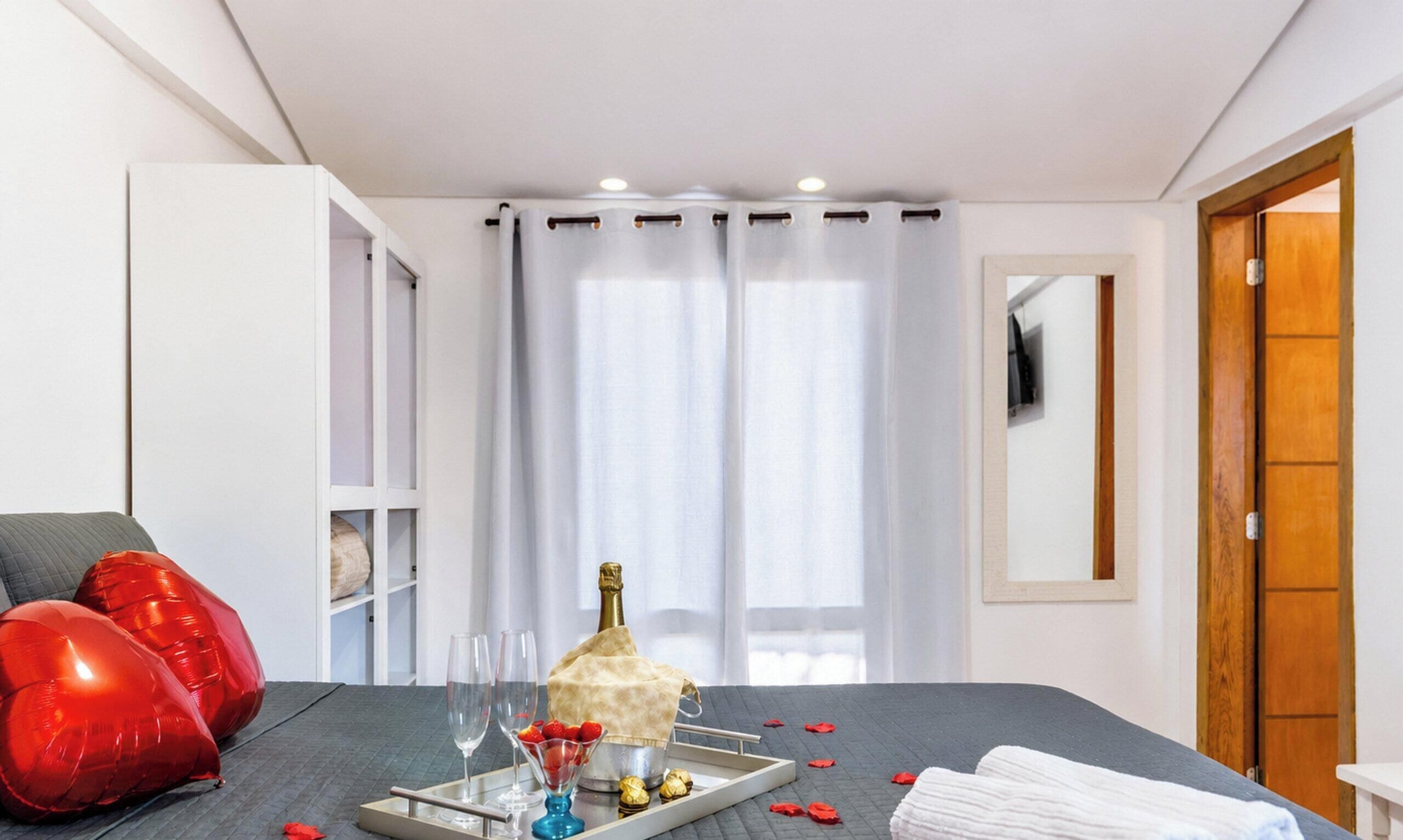 Quarto de hotel com uma cama, uma bandeja com champanhe, copos e chocolates, um balão vermelho e uma porta de madeira.