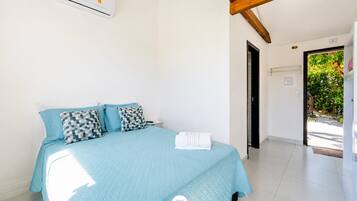 Quarto com cama, almofadas, ar condicionado branco e porta que dá para um jardim.