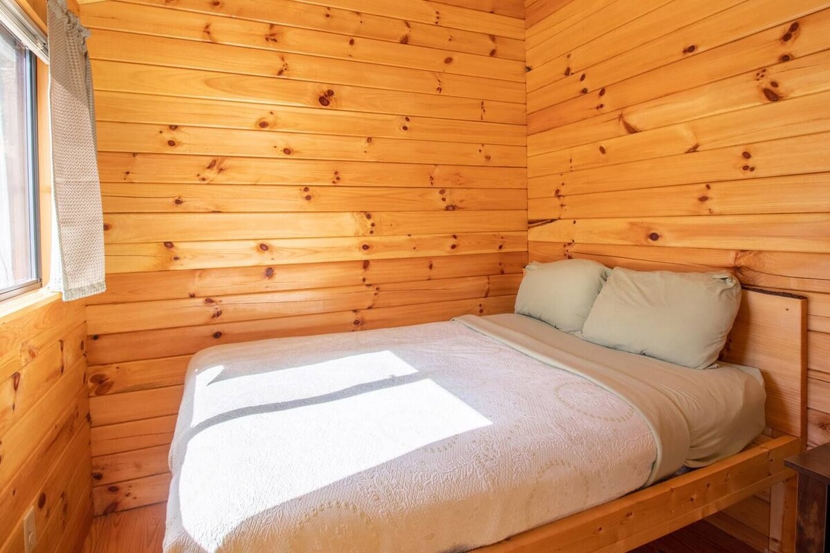 Quarto de cabana de madeira com cama, almofadas e janela com cortina.