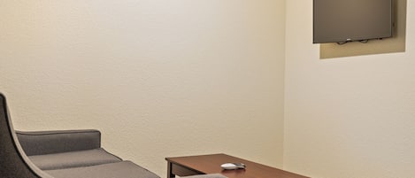 Um quarto pequeno e reservado, com uma única cadeira cinza, uma mesa de madeira e uma televisão de tela plana fixada na parede.