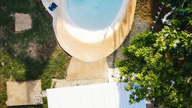 Vista aérea de uma piscina com formato peculiar, rodeada por gramado, algumas plantas e mobiliário de exterior.