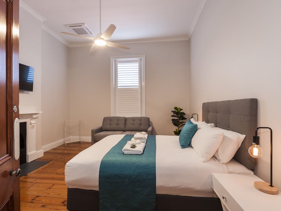Quarto com cama, uma cadeira estofada cinza, ventoinha no teto, televisão e mesa de cabeceira com abajur.