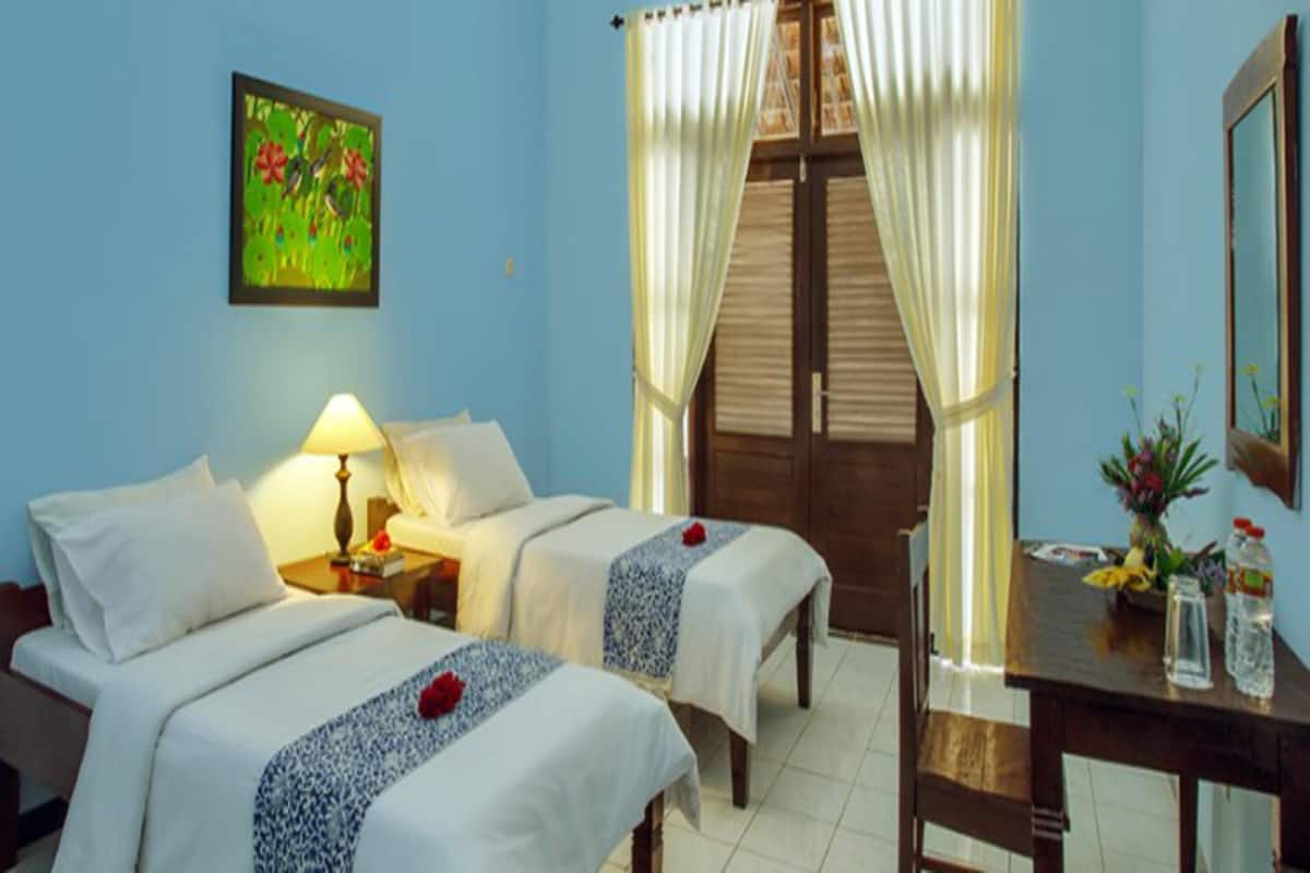Quarto de hotel com duas camas, uma mesa de cabeceira com uma luminária, uma porta de madeira, uma pequena mesa com um vaso e garrafas, e um quadro na parede.